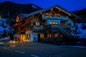 Um hotel com vários andares, com uma varanda no segundo andar, uma escadaria à direita e uma placa com a inscrição "AlpinHotel".
