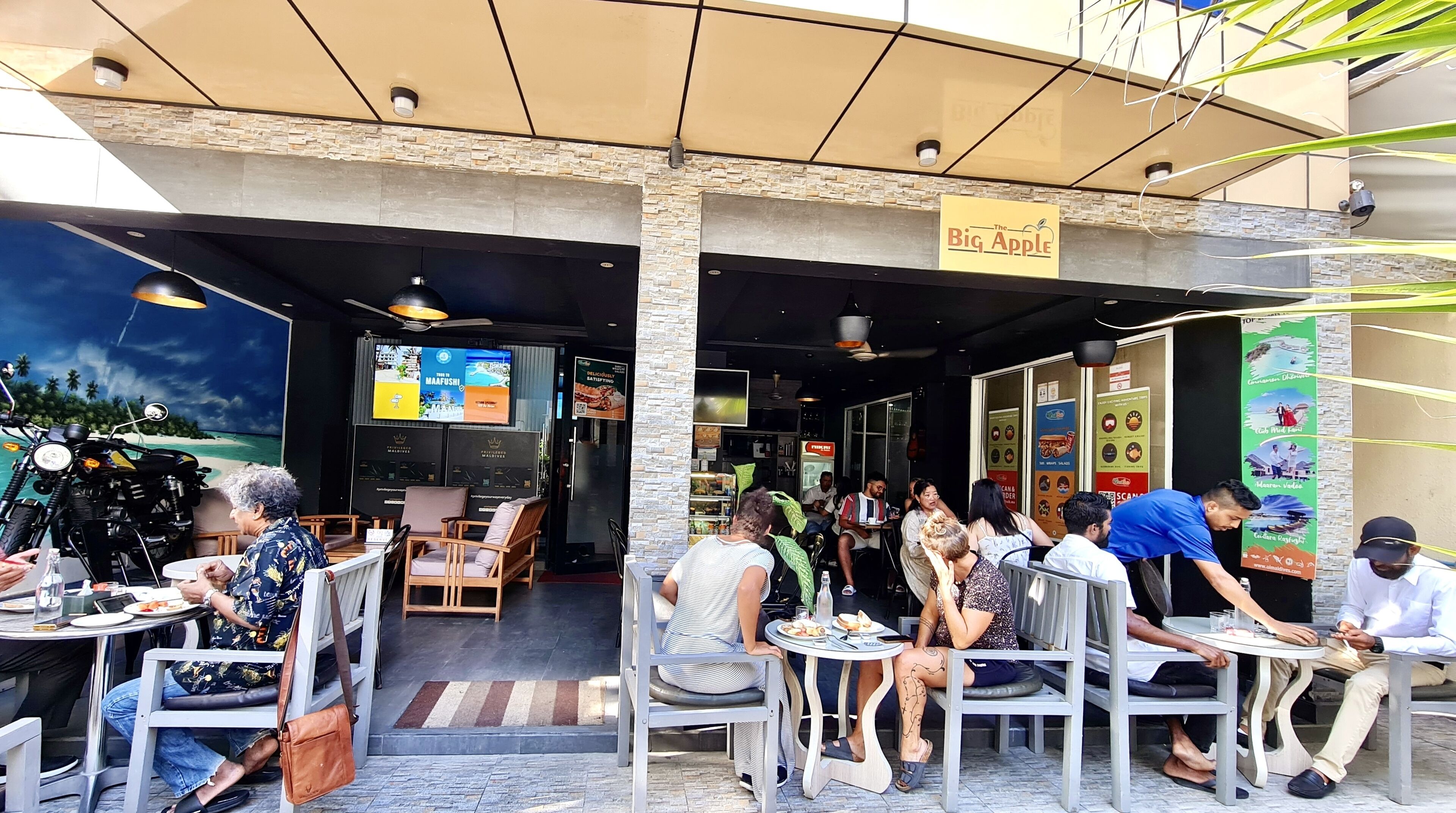 Uma área externa com mesas e cadeiras de um restaurante, um letreiro com 'The Big Apple' e um mural de uma paisagem tropical.