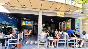 Uma área externa com mesas e cadeiras de um restaurante, um letreiro com 'The Big Apple' e um mural de uma paisagem tropical.