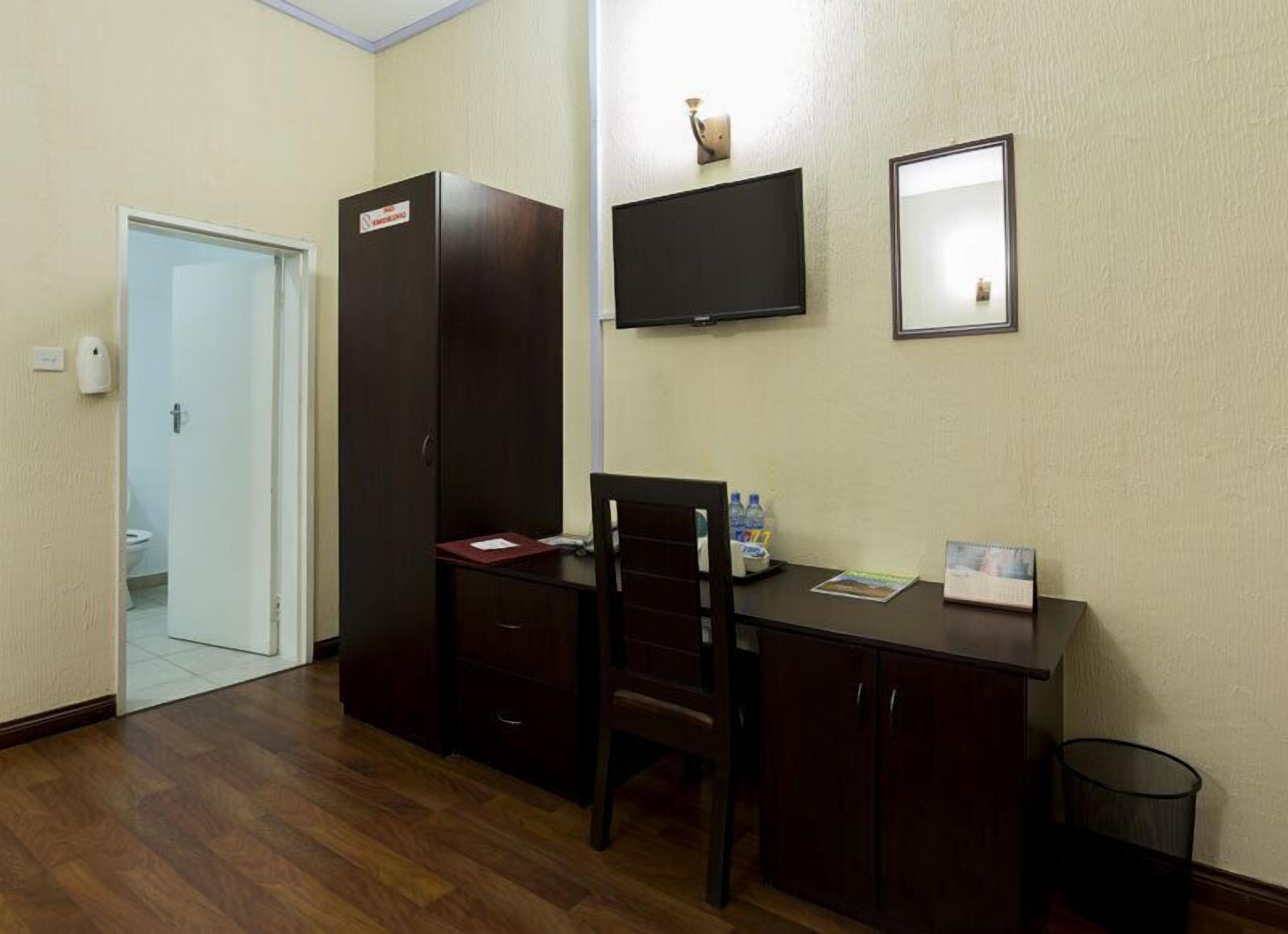 Quarto de hotel com escrivaninha de madeira, uma cadeira, televisão de tela plana na parede, prateleira pequena com um vaso e uma lixeira.