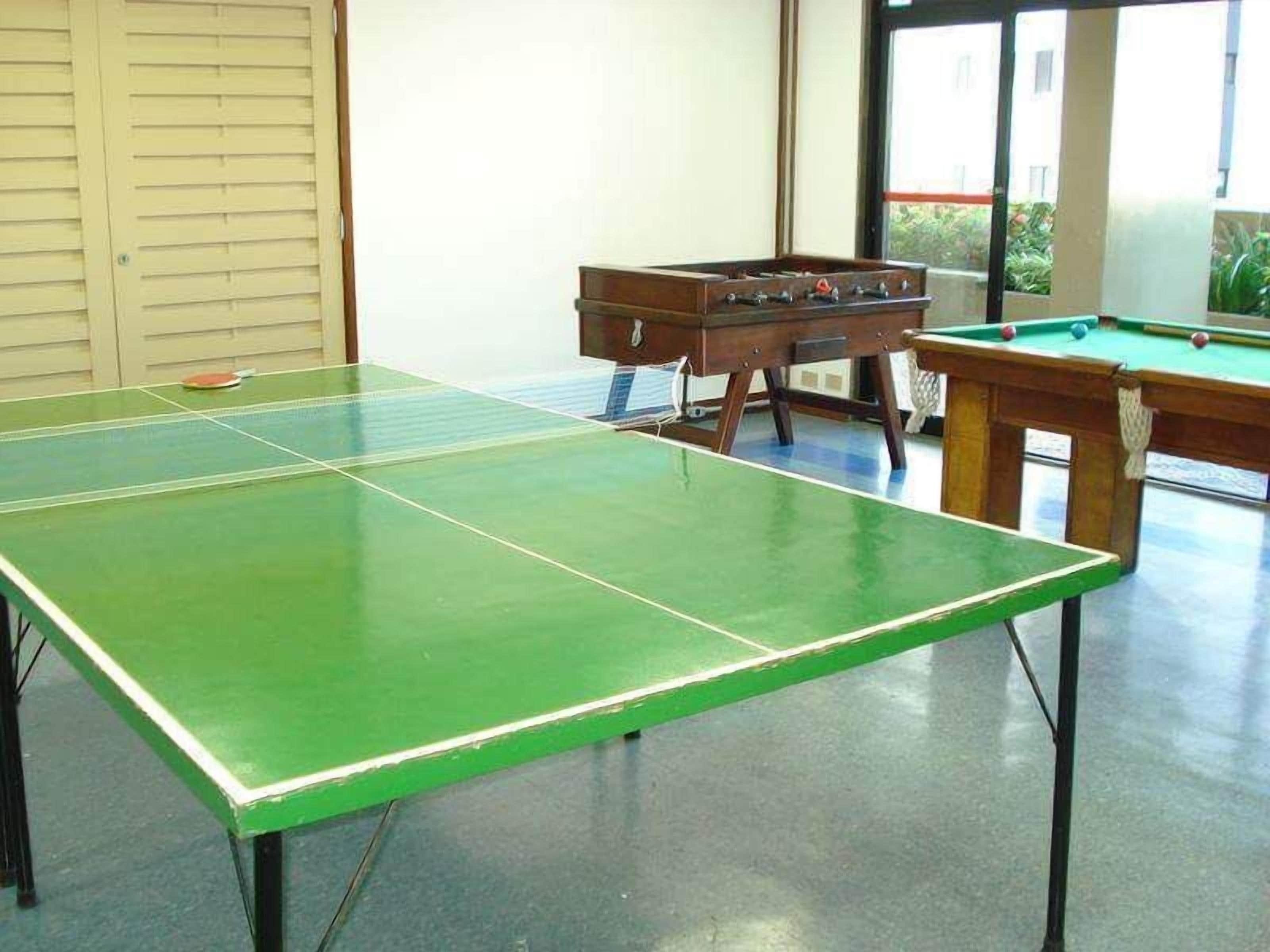 Uma mesa de bilhar com superfície de feltro verde, uma mesa de sinuca e uma mesa de futebol de mesa em um quarto com uma janela grande.