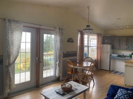 Uma área de refeições com mesa e cadeiras, uma cozinha com armários e uma geladeira, e uma vista pela janela.