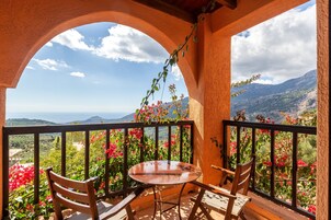 Varanda com mesa redonda e cadeiras, cercada por guarda-corrimão preto, com vista para uma paisagem montanhosa com flores vermelhas e vegetação.