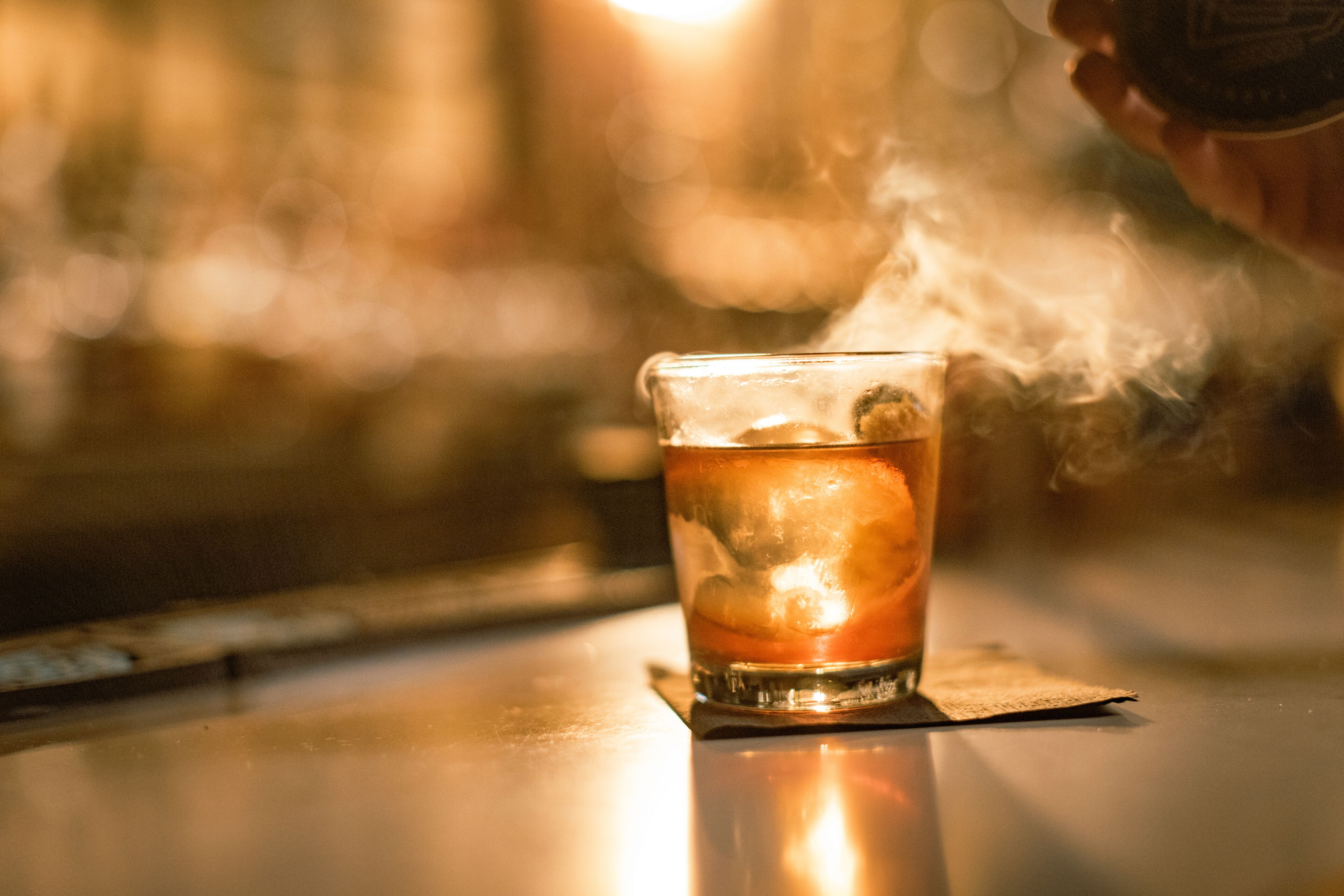 Um copo com líquido âmbar e cubos de gelo sobre uma superfície refletora, possivelmente um balcão de bar, com fundo desfocado que sugere um ambiente interno.