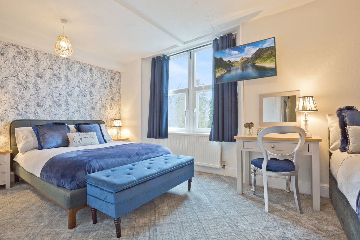 Quarto com cama grande, escrivaninha, cadeira, janela com cortinas e um quadro na parede.