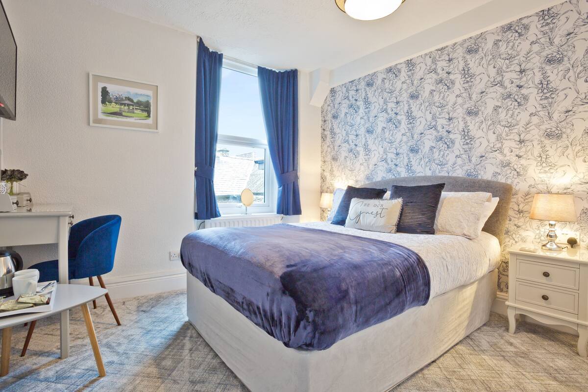 Quarto com cama, escrivaninha, cadeira e janela com cortinas.