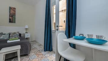 Quarto de hotel pequeno com cama, uma mesinha com uma tigela, uma cadeira e uma janela com cortinas.