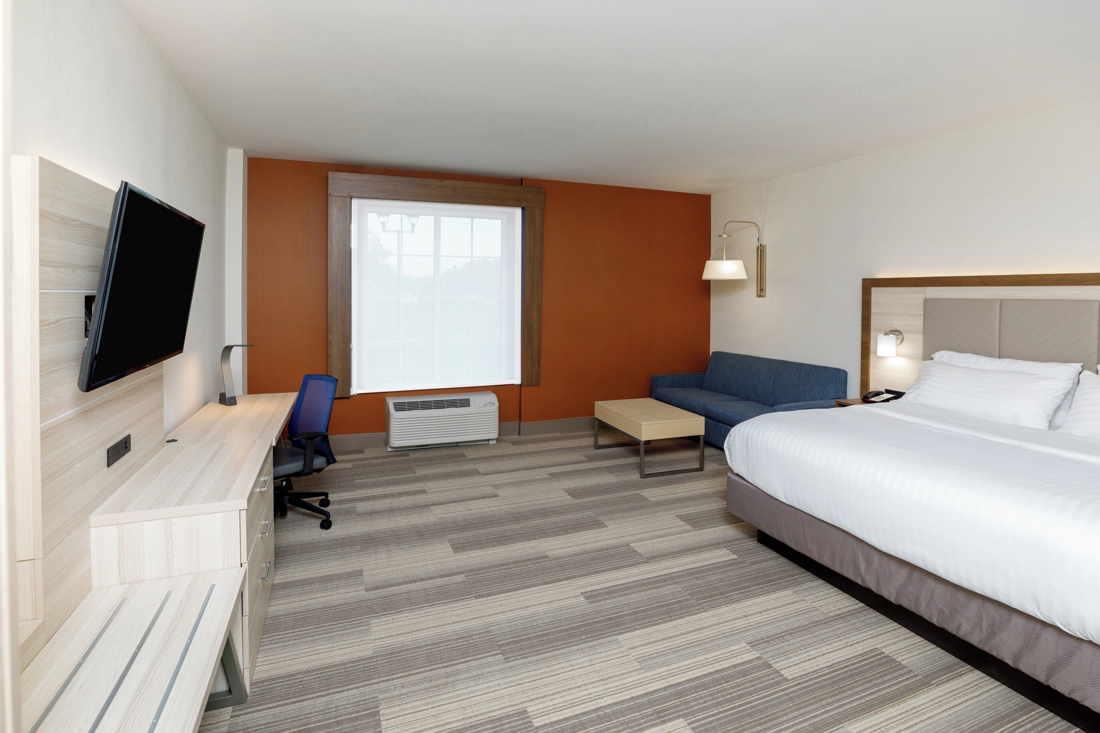 Quarto de hotel com cama, escrivaninha, televisão de tela plana, cadeira azul, mesinha e janela.