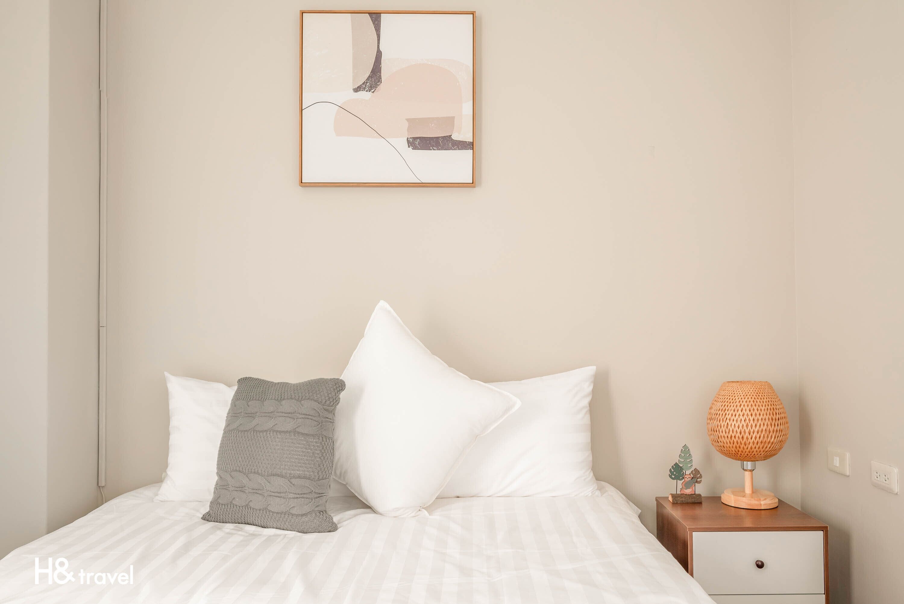 Cama bem arrumada com roupa de cama branca e um travesseiro de lã cinza, uma pequena mesa de cabeceira de madeira com luminária e uma obra de arte abstrata emoldurada na parede.
