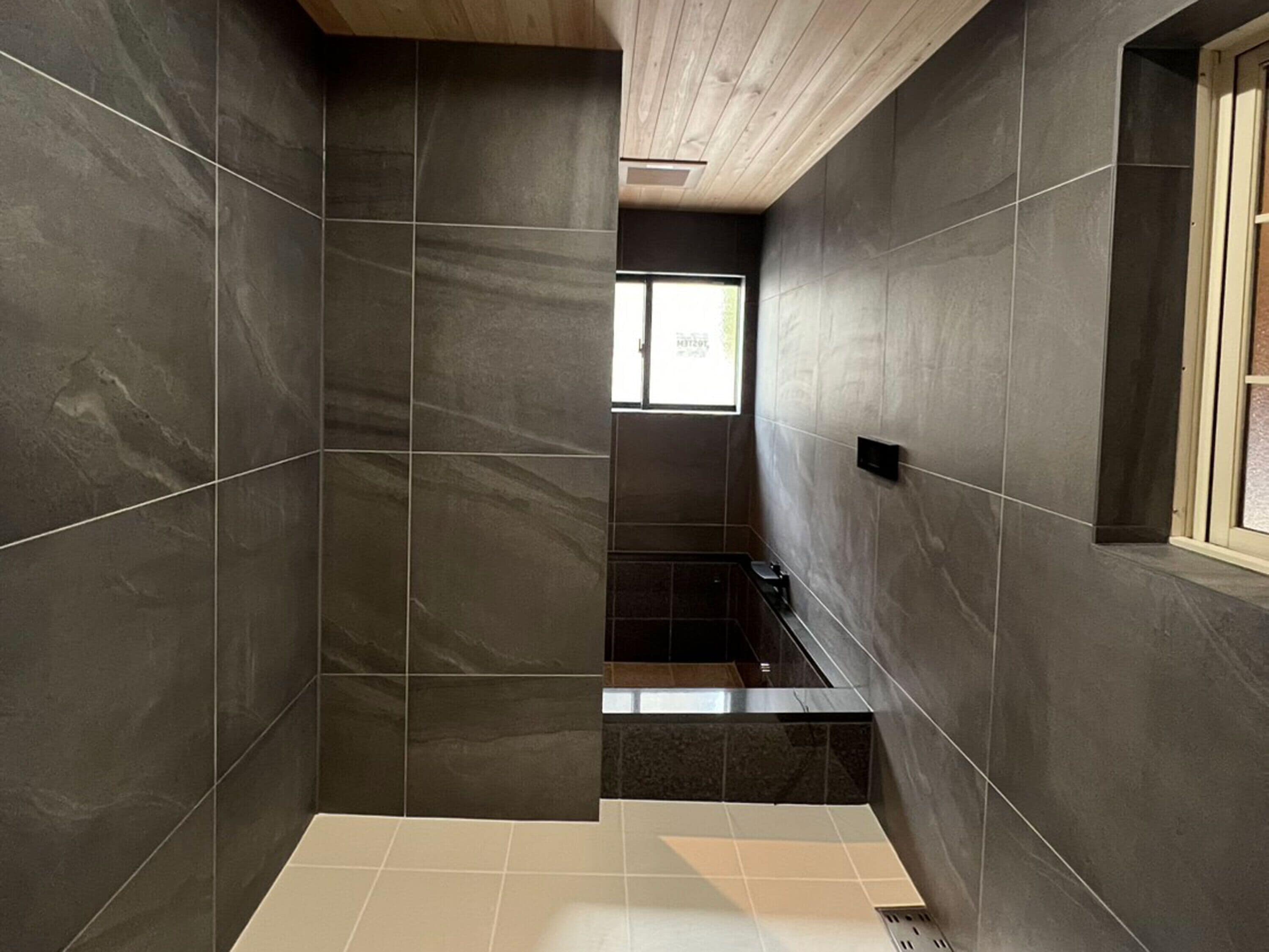 Um banheiro moderno com azulejos escuros, teto de madeira e janela que permite iluminação natural.