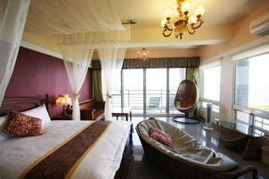 Um quarto espaçoso com uma cama grande, um chaise-lounge, um espaço de estar com uma poltrona e vista para o exterior por grandes janelas.