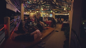 Espaço interior com mesas e cadeiras dispostas para refeições, área de bar e zona de estar com almofadas.