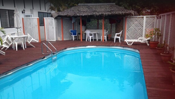 Uma piscina com deck de madeira, espreguiçadeiras brancas e área de estar coberta.