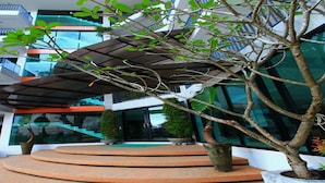 Edifício moderno com escadaria curva que leva a uma entrada coberta, cercado por plantas em vasos e fachada de vidro.
