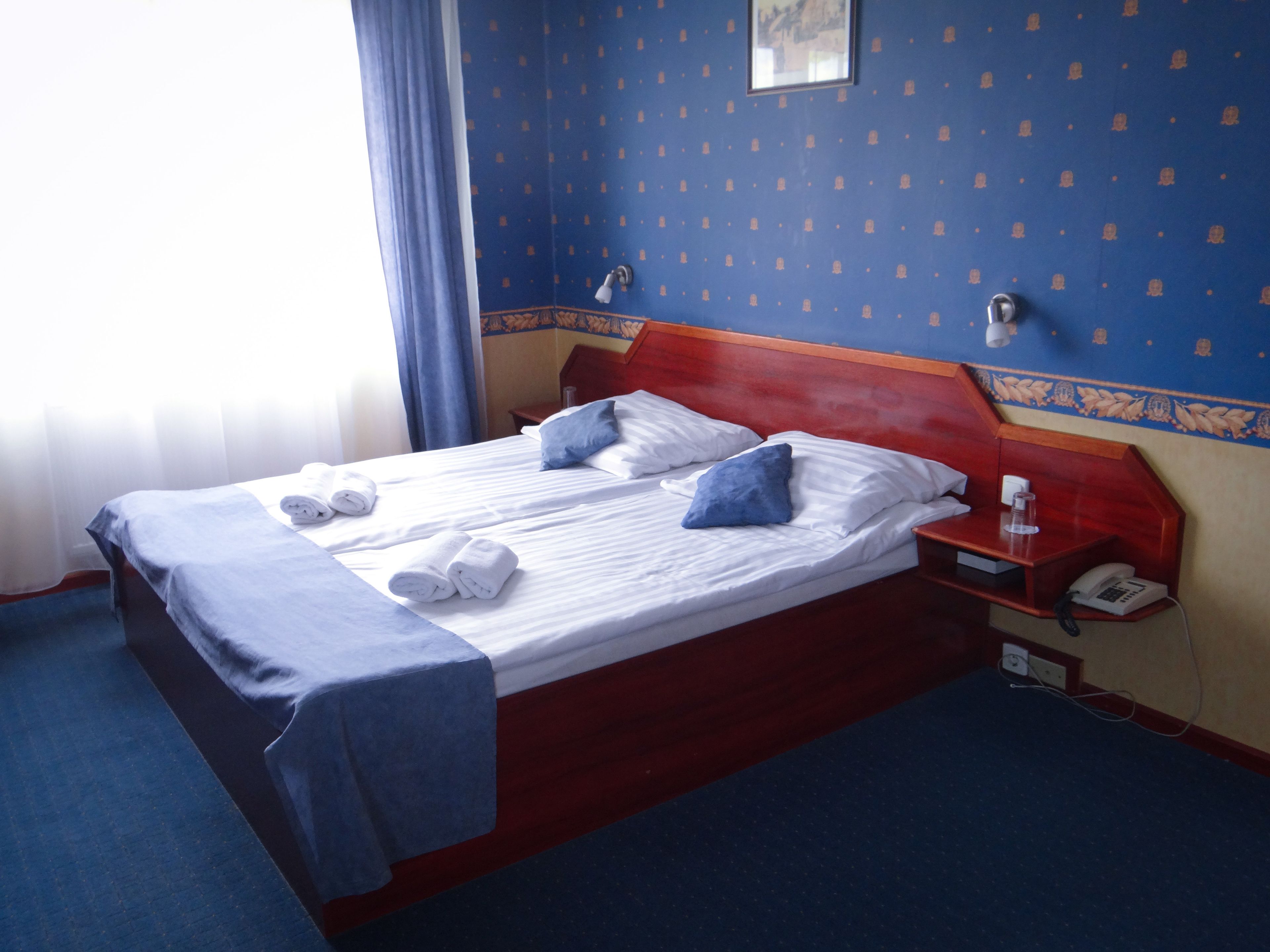 Uma cama de casal com lençóis brancos e almofadas azuis, uma cabeceira de madeira, uma mesinha de cabeceira com telefone e uma parede com design estampado.