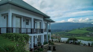Um edifício branco de dois andares com varanda, rodeado por vegetação e vista para um lago e montanhas.