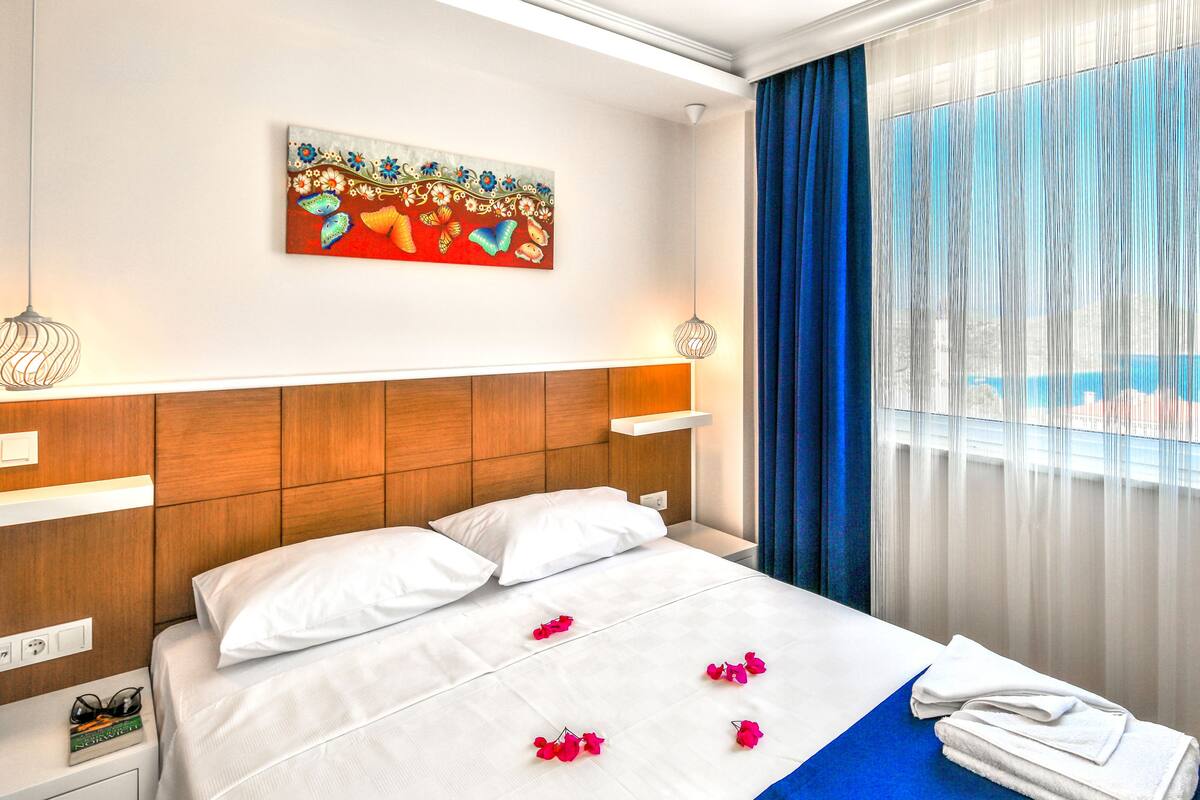Quarto de hotel com cabeceira de madeira, roupa de cama branca, um quadro decorativo, uma janela com cortinas azuis e uma cama bem arrumada com toalhas.
