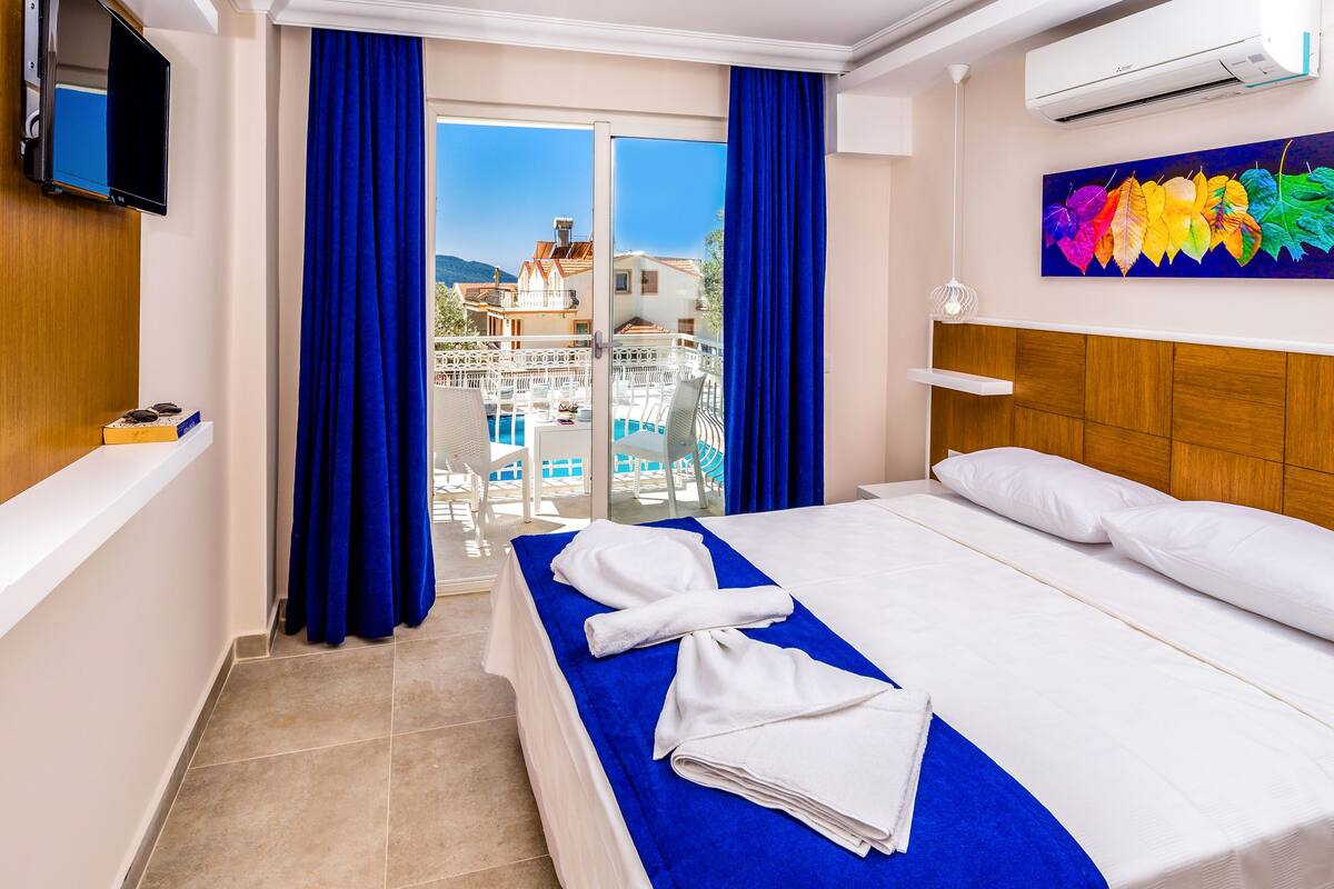 Quarto de hotel com cama, televisão, varanda com piscina e um quadro de folha colorida na parede.