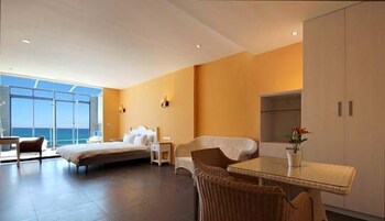 Quarto de hotel moderno com uma cama grande, área de estar com mesa de vidro e vista para o oceano.