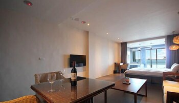 Quarto de hotel com cama, mesa de jantar com copos de vinho, televisão fixada na parede e vista para o mar através de amplas janelas.