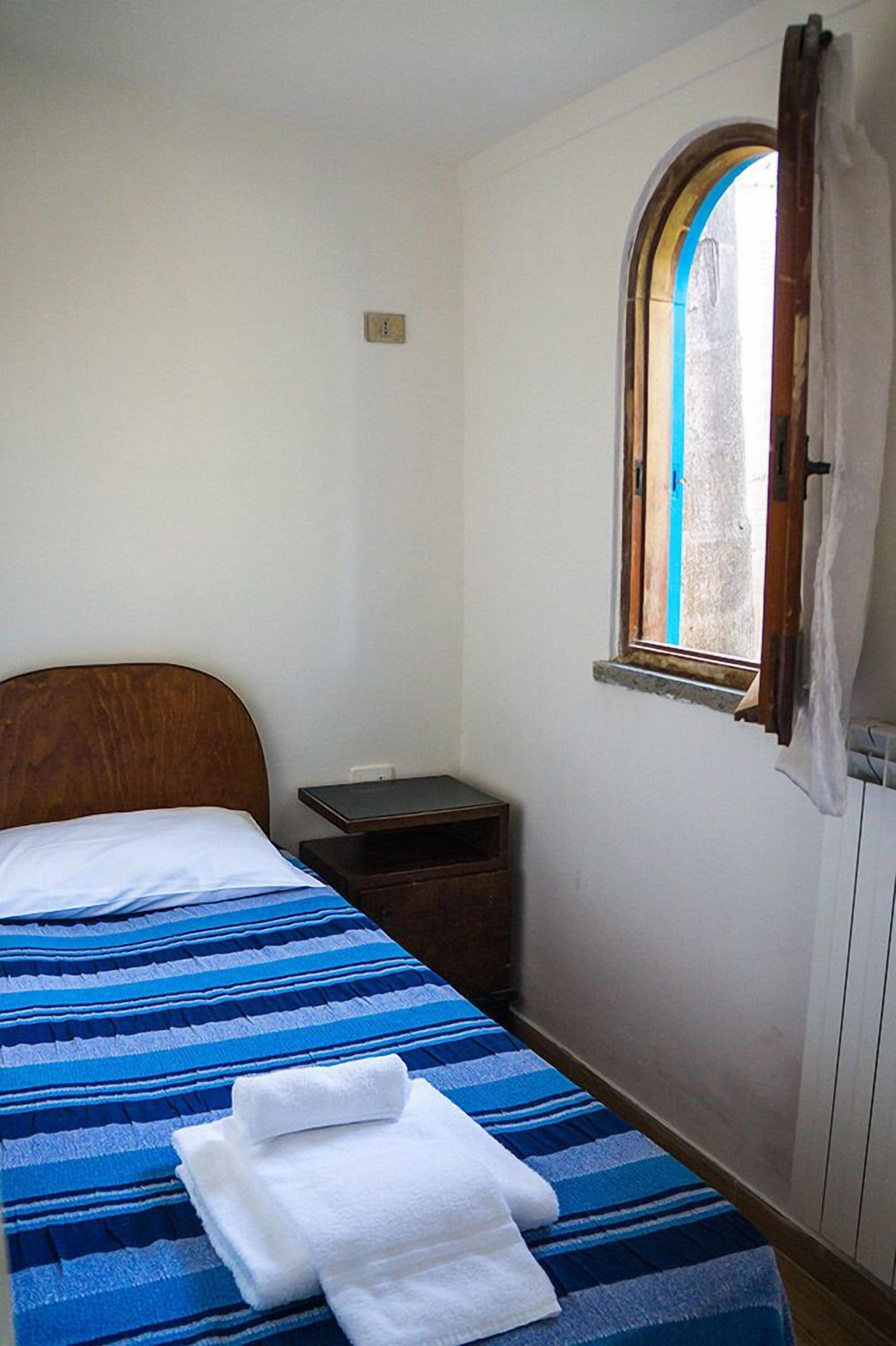 Quarto pequeno com uma cama de solteiro, coberta por um edredom listrado azul e branco, uma cabeceira de madeira, uma mesinha de cabeceira, uma janela com moldura de madeira e detalhes azuis, e uma toalha branca sobre a cama.