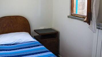 Quarto pequeno com uma cama de solteiro, coberta por um edredom listrado azul e branco, uma cabeceira de madeira, uma mesinha de cabeceira, uma janela com moldura de madeira e detalhes azuis, e uma toalha branca sobre a cama.