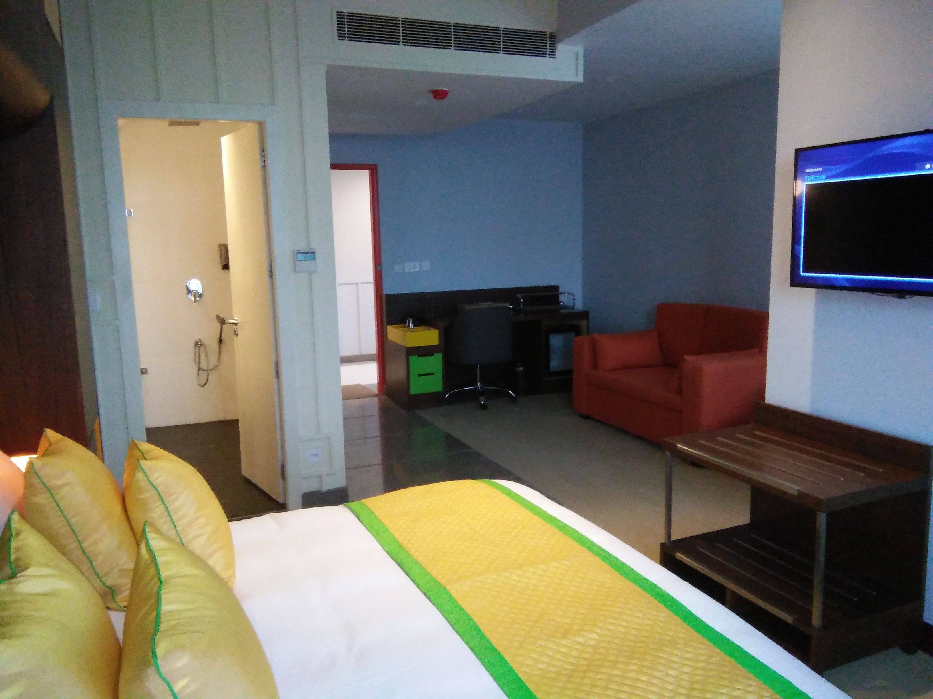 Quarto de hotel com uma cama, uma escrivaninha, uma cadeira vermelha e uma televisão na parede.