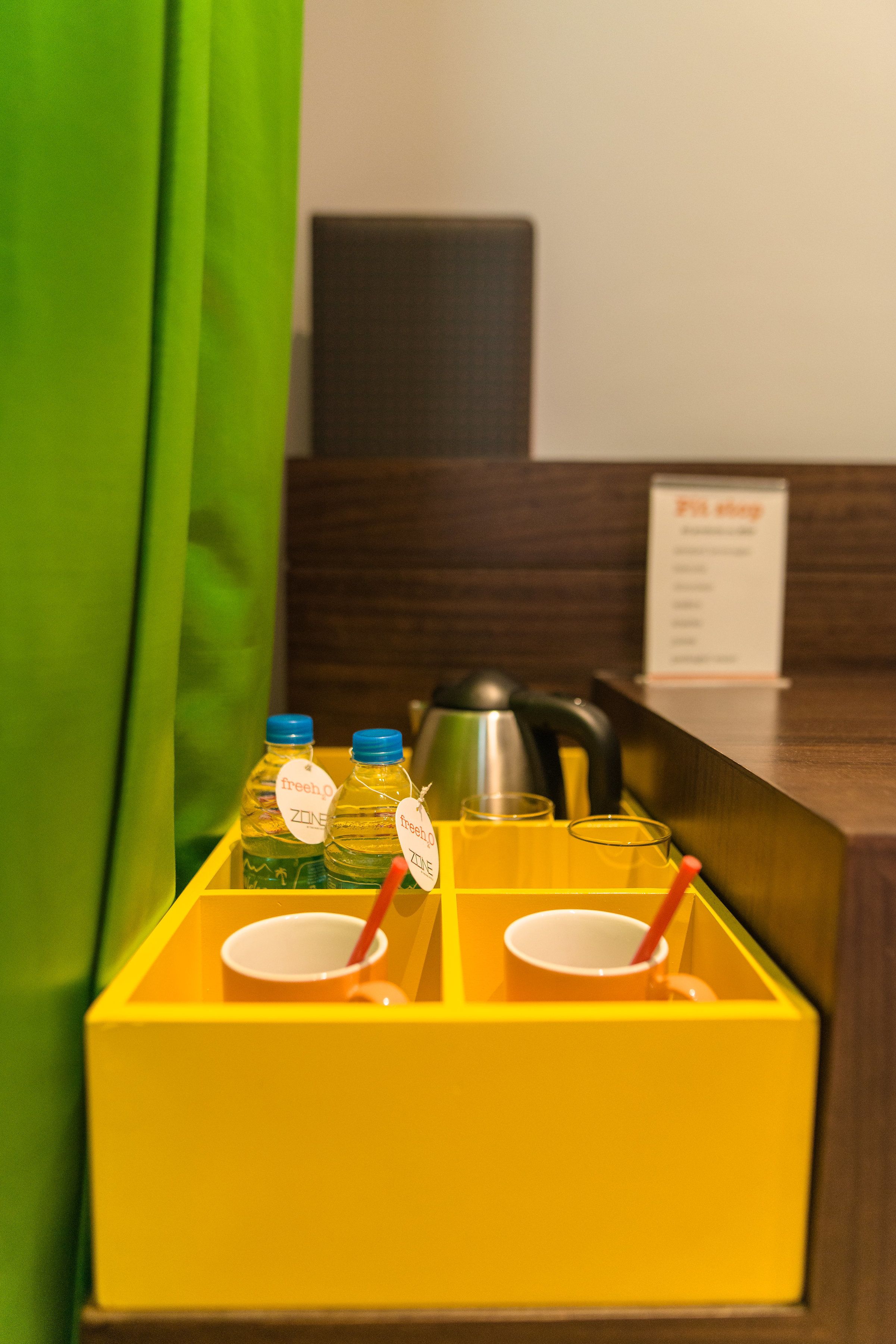 Uma área de recepção de hotel com uma bandeja amarela contendo duas garrafas de água, duas copos com canudos e uma chaleira.