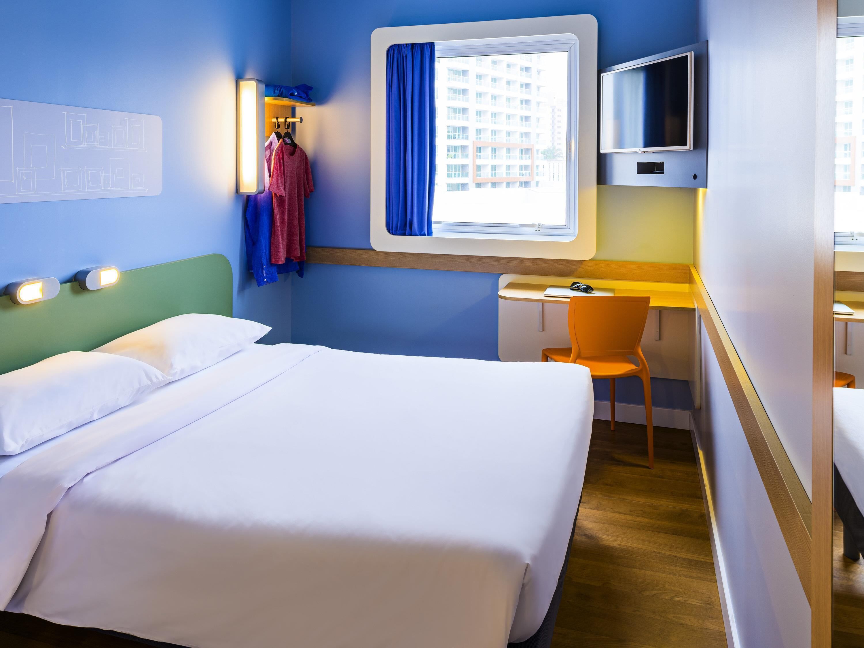 Quarto de hotel com cama, escrivaninha, cadeira, televisão e janela com vista para edifícios.