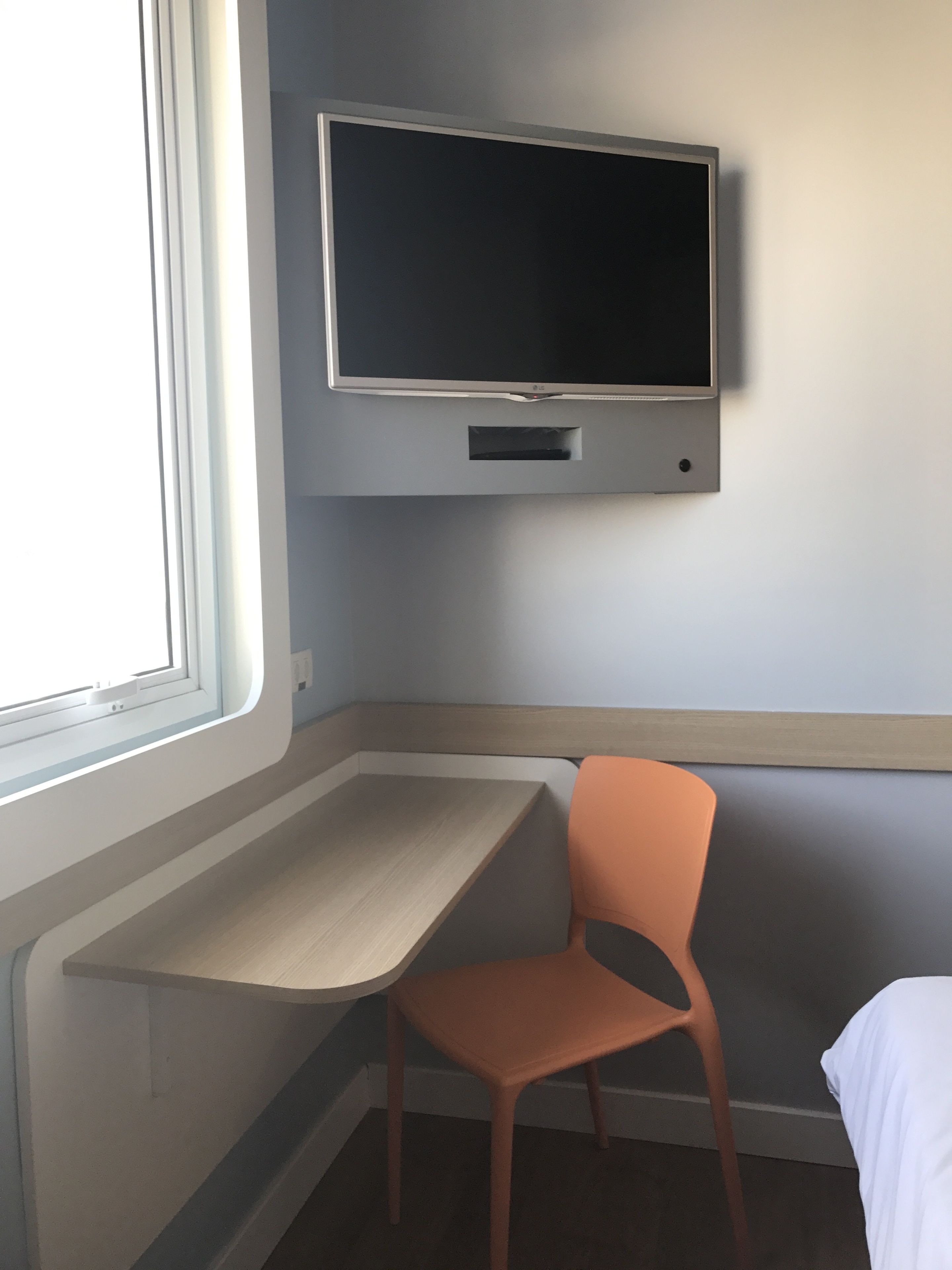 Quarto de canto com televisão na parede, escrivaninha de madeira, cadeira laranja e cama com roupa de cama branca.