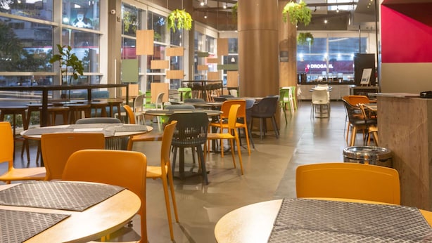 Interior moderno de café com mesas, cadeiras, plantas pendentes e janelas amplas.