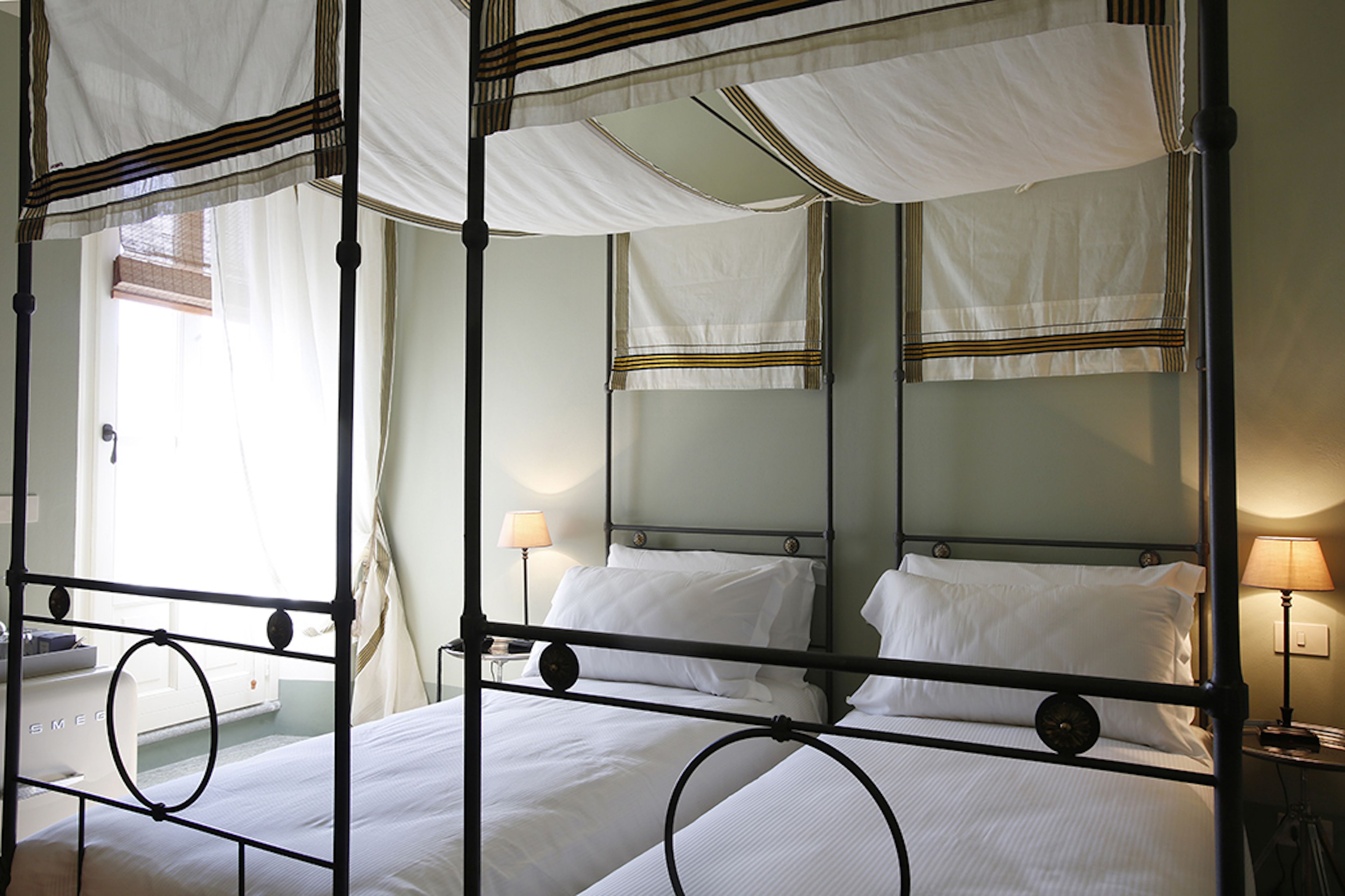 Uma cama de dossel com lençóis brancos, uma mesinha de cabeceira com abajur e uma janela com cortinas.