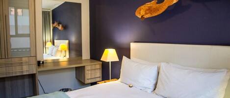 Quarto de hotel com cama, escrivaninha, espelho e uma escultura de peixe na parede.
