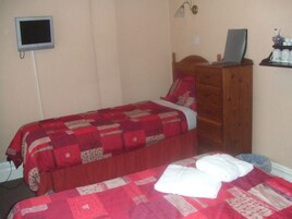 Quarto com cama, cômoda de madeira, escrivaninha com computador e televisão na parede.