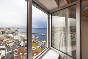 Quarto em um arranha-céu com uma grande janela que oferece vista para a paisagem costeira urbana, incluindo o mar e edifícios.