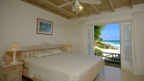 Um quarto com cama, duas mesas de cabeceira, um ventilador de teto, um quadro e vista para a praia.