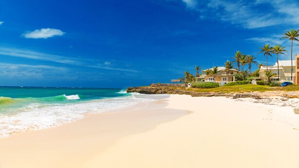 Uma praia com águas turquesa cristalinas, areia branca e algumas palmeiras. Edifícios com estilo arquitetônico tropical podem ser vistos ao fundo.