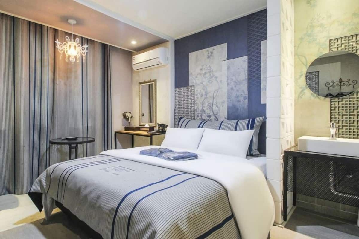 Quarto de hotel com uma cama, uma escrivaninha, uma cadeira, uma mesa redonda pequena, um espelho e um banheiro com pia e chuveiro.