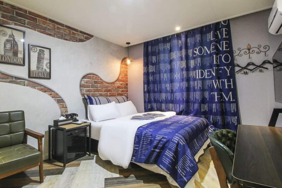 Quarto com parede de tijolos, cama com colcha azul e branca, uma cadeira de madeira, uma pequena mesa de cabeceira e um cartaz na parede com a inscrição 'Live Someone Identity With Them'.