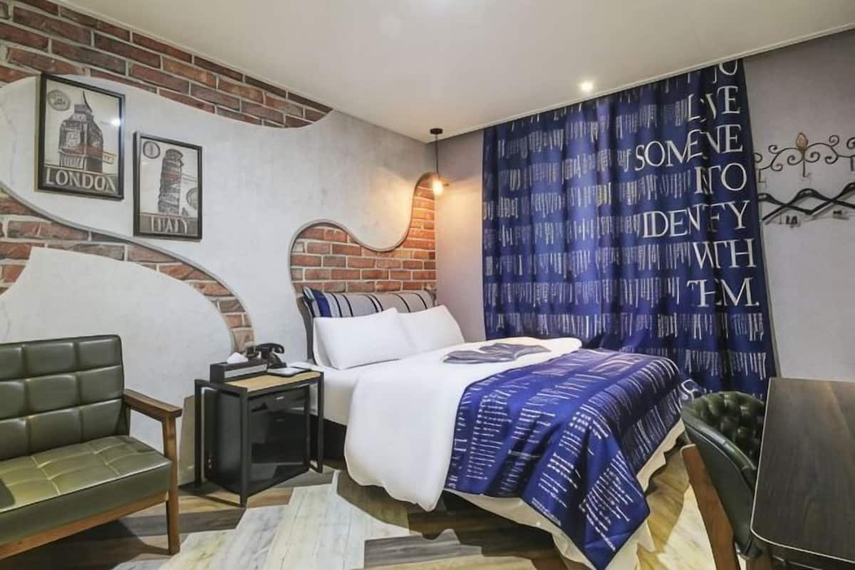 Um quarto com parede de tijolos, uma cama com colcha azul e branca, cabeceira, mesa de cabeceira e uma poltrona verde.