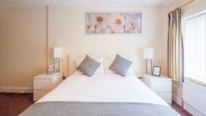 Quarto de hotel com uma cama grande, duas mesas de cabeceira, um criado-mudo, um quadro na parede e uma janela com cortinas.