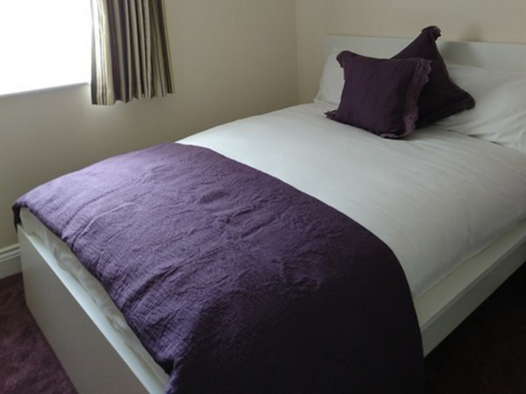 Quarto individual com uma cama de solteiro, cabeceira branca, manta roxa e travesseiro branco.