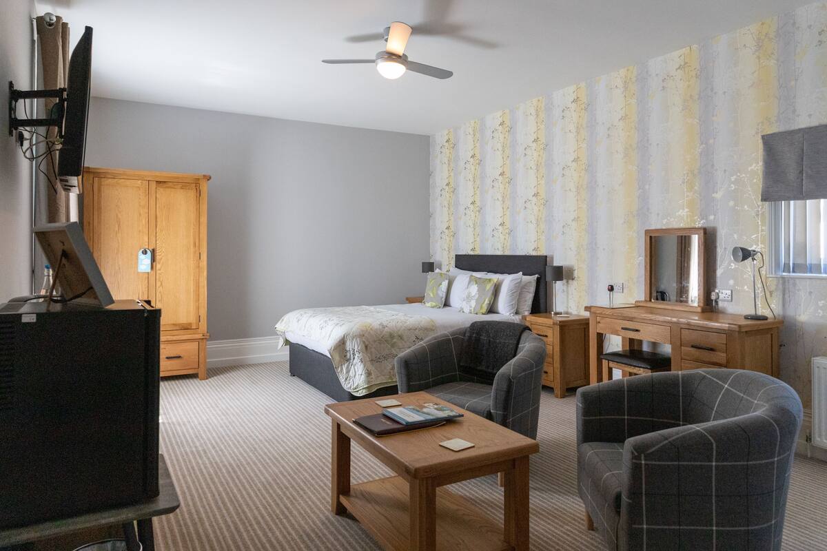 Quarto com cama, duas poltronas, escrivaninha de madeira, televisão e ventoinha de teto.