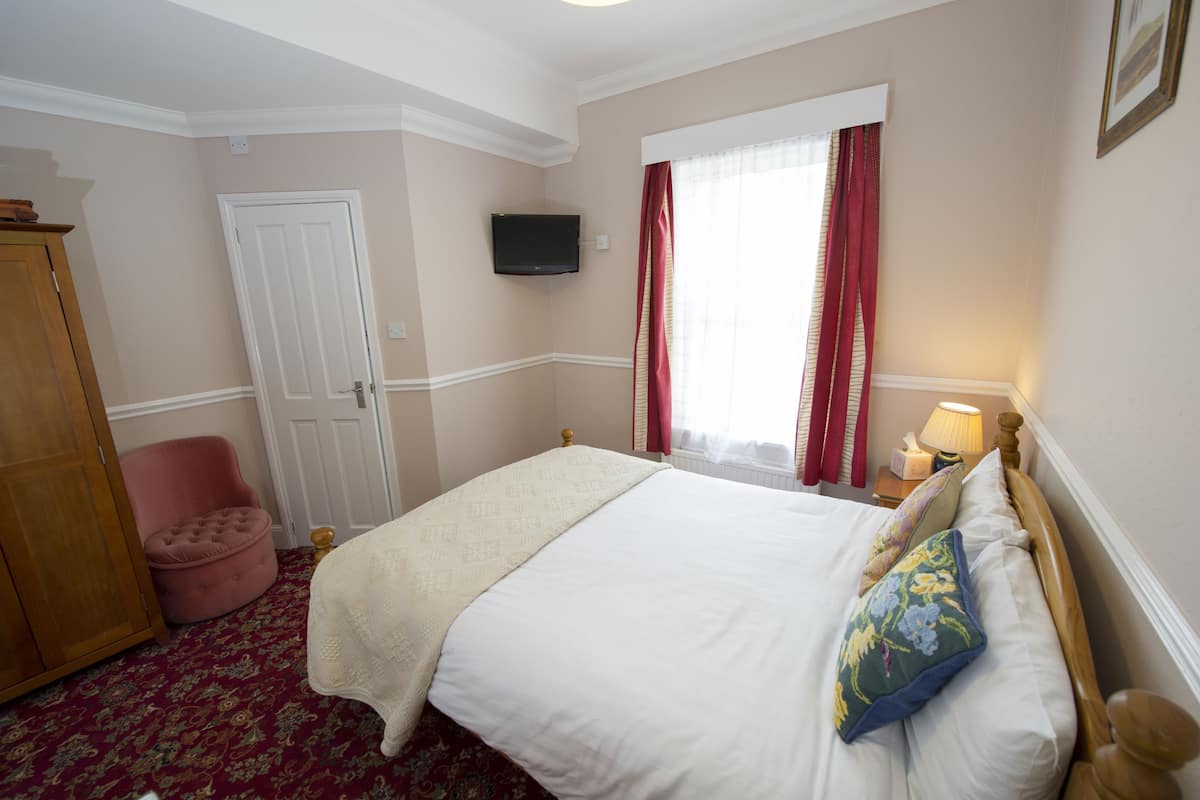 Quarto com cama, televisão na parede, janela com cortinas, guarda-roupa de madeira e uma pequena cadeira rosa.