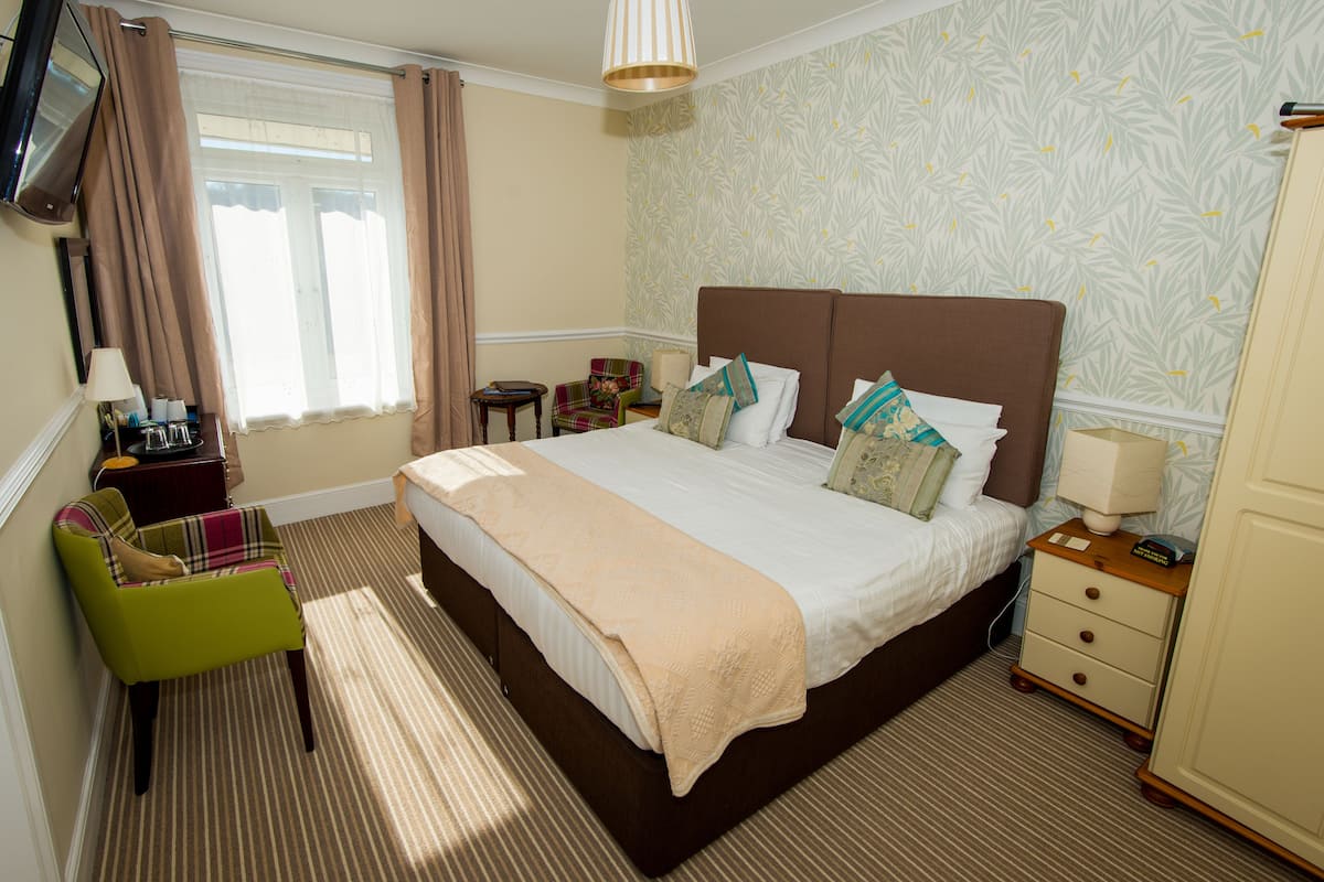 Quarto de hotel bem arrumado com uma cama grande, uma poltrona verde, uma mesinha de cabeceira com abajur e um aparador.