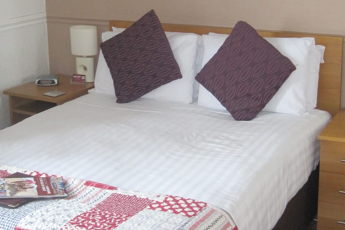 Cama bem arrumada com colcha estampada, duas almofadas, mesa de cabeceira com abajur e uma obra de arte floral na parede.