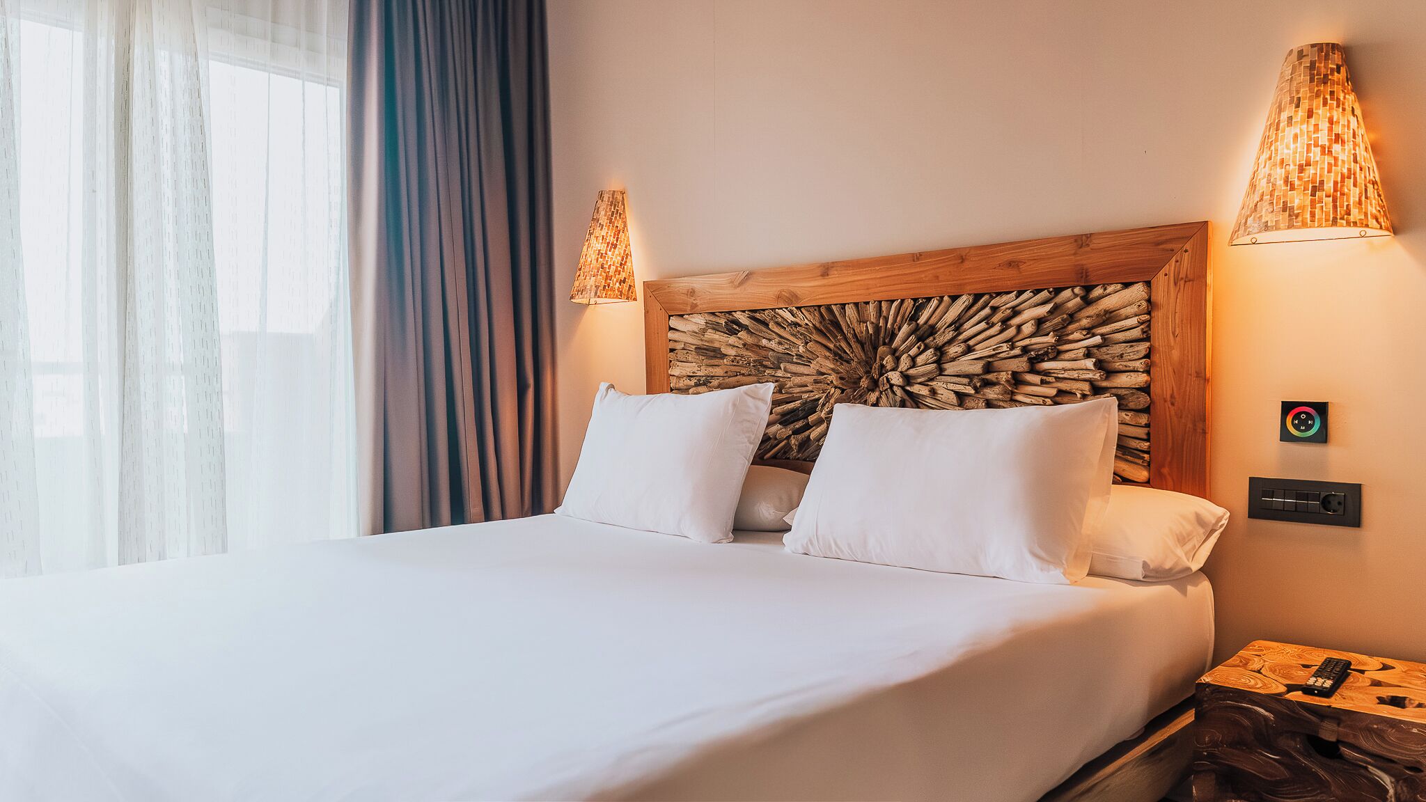 Quarto de hotel com cabeceira de madeira, roupa de cama branca, mesa de cabeceira com controle remoto e luminária fixada na parede.