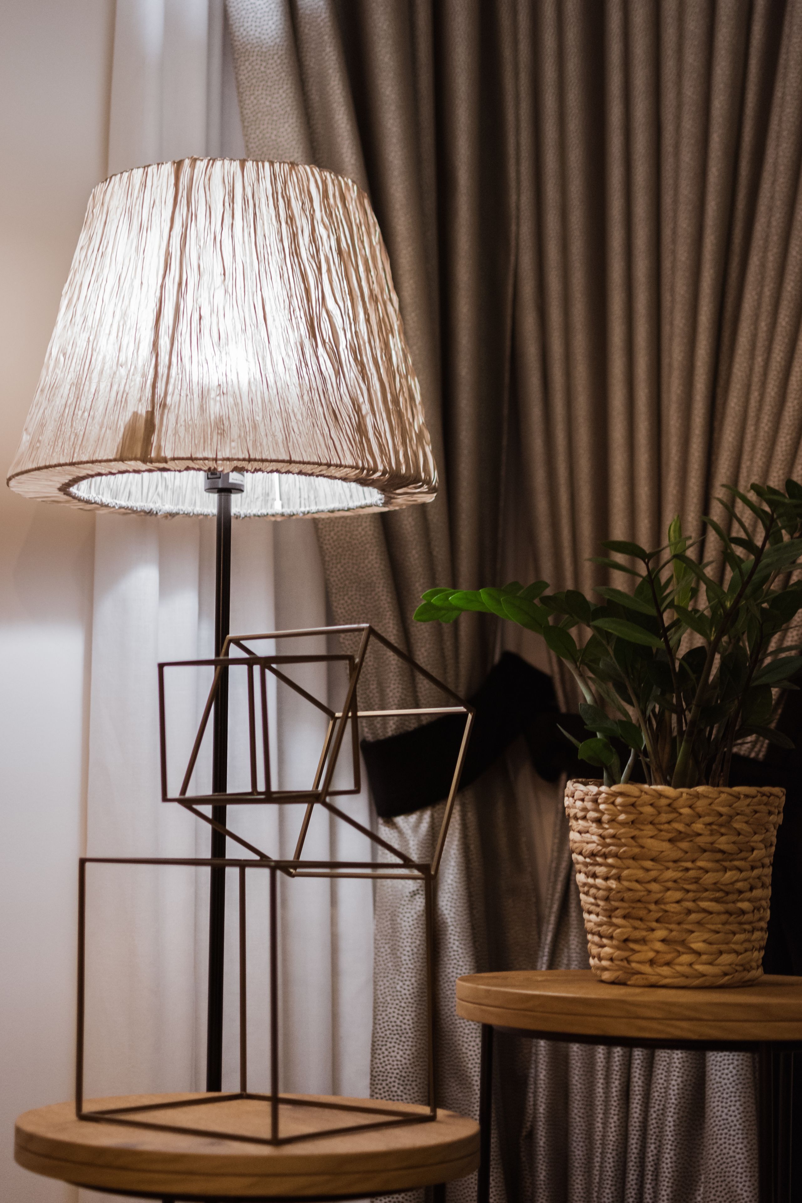 Um abajur com cúpula trançada, base de metal geométrica e uma planta em vaso sobre uma mesa redonda de madeira.