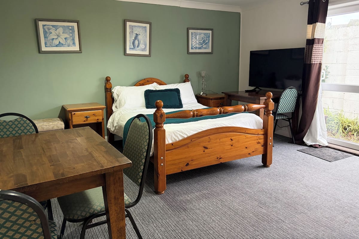 Quarto com cama de madeira, uma escrivaninha, uma cadeira e uma televisão.