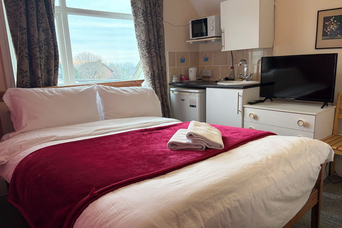Quarto de hotel com cama, manta vermelha, televisão, micro-ondas e pia.